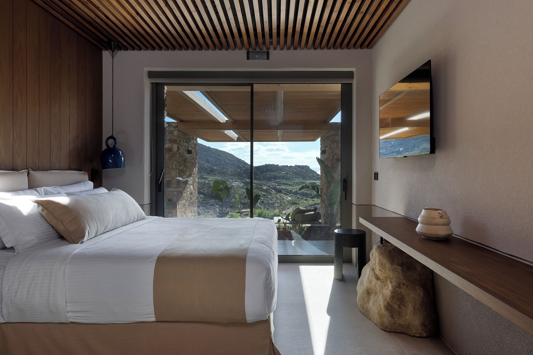 Quarto com uma cama grande, parede de pedra, teto de madeira, televisão e vista para montanhas.