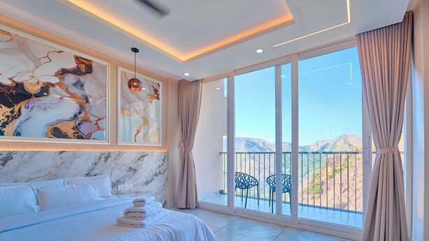 Quarto com cama grande, parede de mármore, varanda com cadeiras e vista para a montanha.