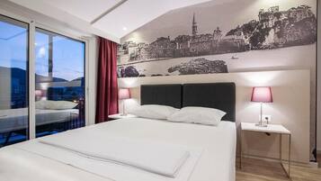 Quarto de hotel com uma cama grande, duas mesas de cabeceira com luminárias, uma porta de vidro deslizante para uma varanda e um mural com vista urbana na parede.