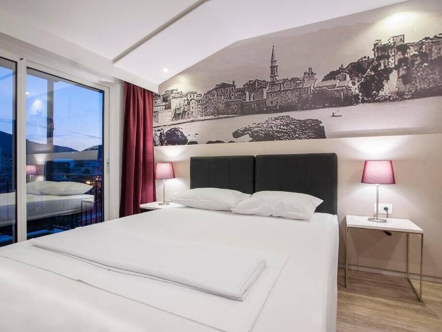 Quarto de hotel com uma cama grande, duas mesas de cabeceira com luminárias, uma porta de vidro deslizante para uma varanda e um mural com vista urbana na parede.