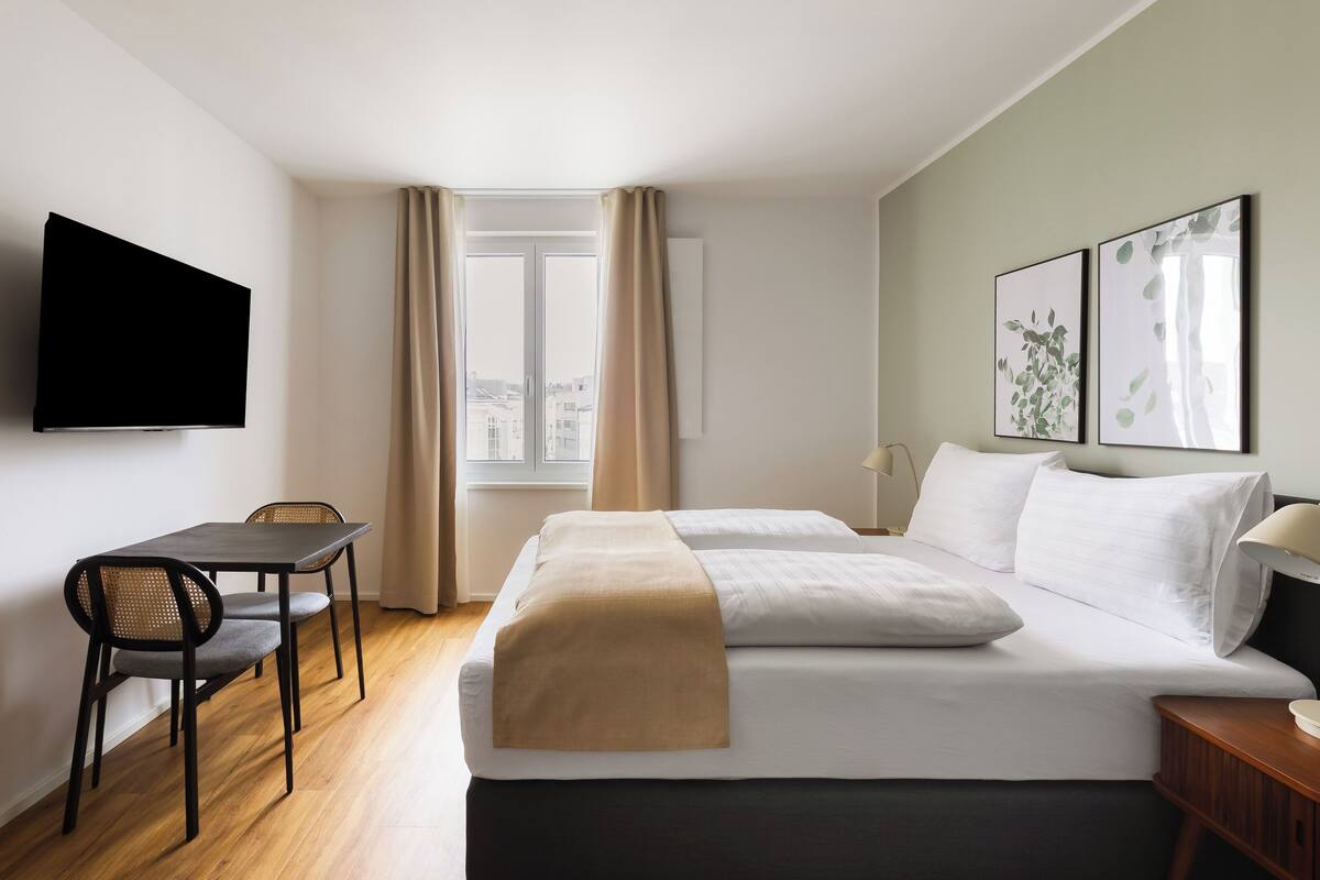 Quarto de hotel com cama, escrivaninha, cadeira, televisão e obras de arte na parede.