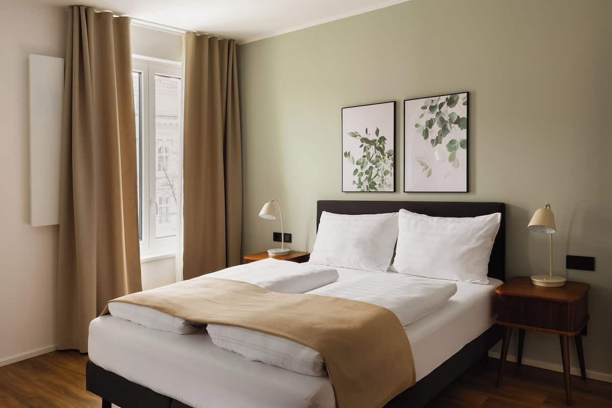 Uma cama bem arrumada com roupa de cama branca e bege, duas luminárias de cabeceira e duas obras de arte com folhas emolduradas na parede.