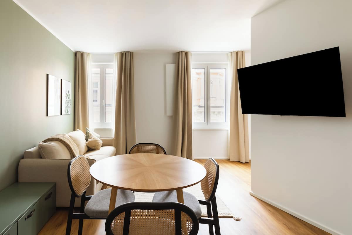 Uma sala de estar moderna com sofá, mesa de jantar redonda e cadeiras, TV na parede e janelas amplas com cortinas.