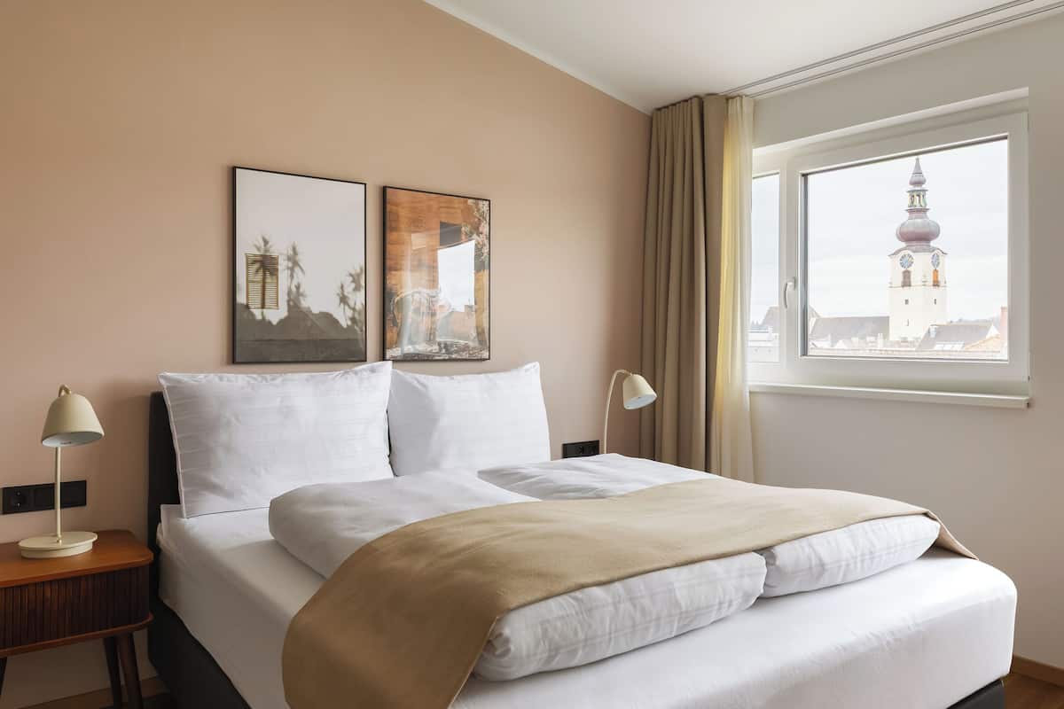 Quarto de hotel com uma cama, duas luminárias de cabeceira, uma janela com vista para uma torre e duas imagens emolduradas na parede.