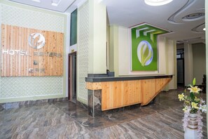 Uma área de recepção de hotel com um balcão de madeira, uma placa na parede e um painel decorativo verde.