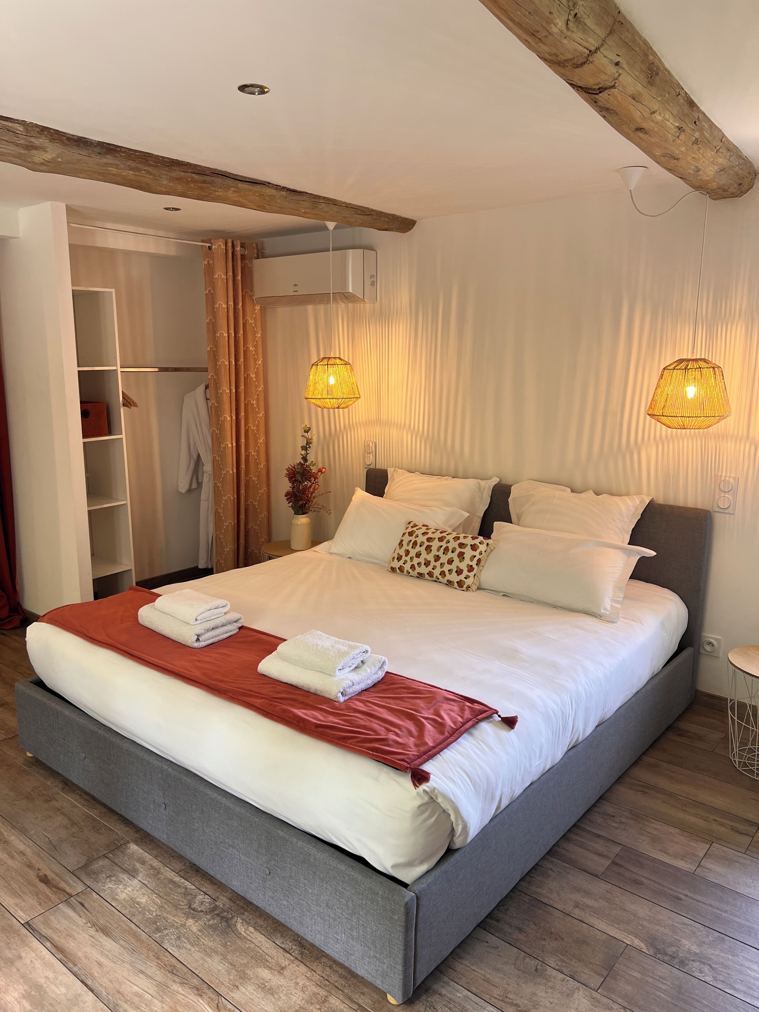 Cama bem arrumada com lençóis brancos e manta vermelha, vigas de madeira no teto, ar condicionado fixado na parede e luminária pendente.