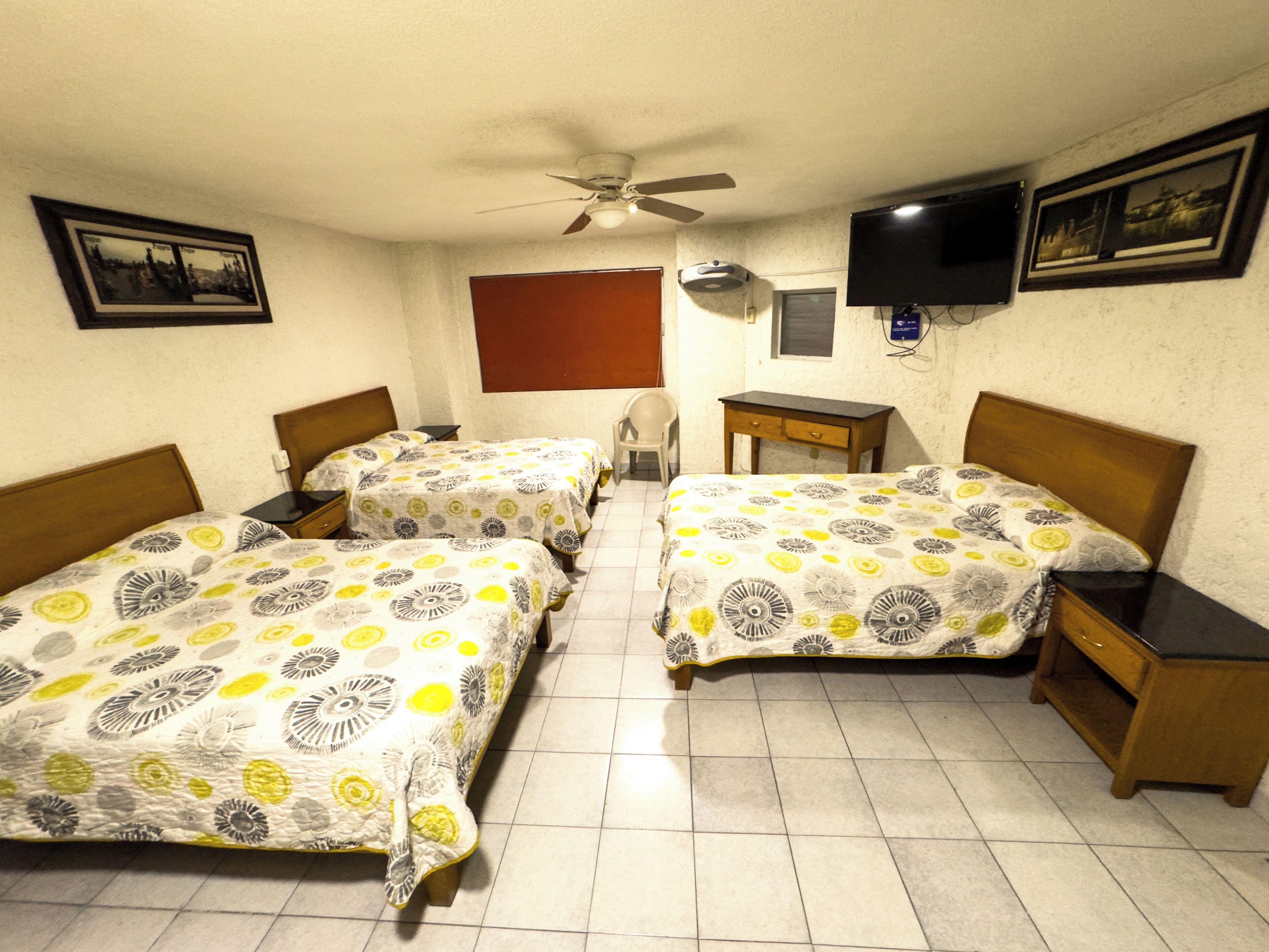 Quarto de hotel com duas camas, ventilador de teto, televisão e uma escrivaninha.