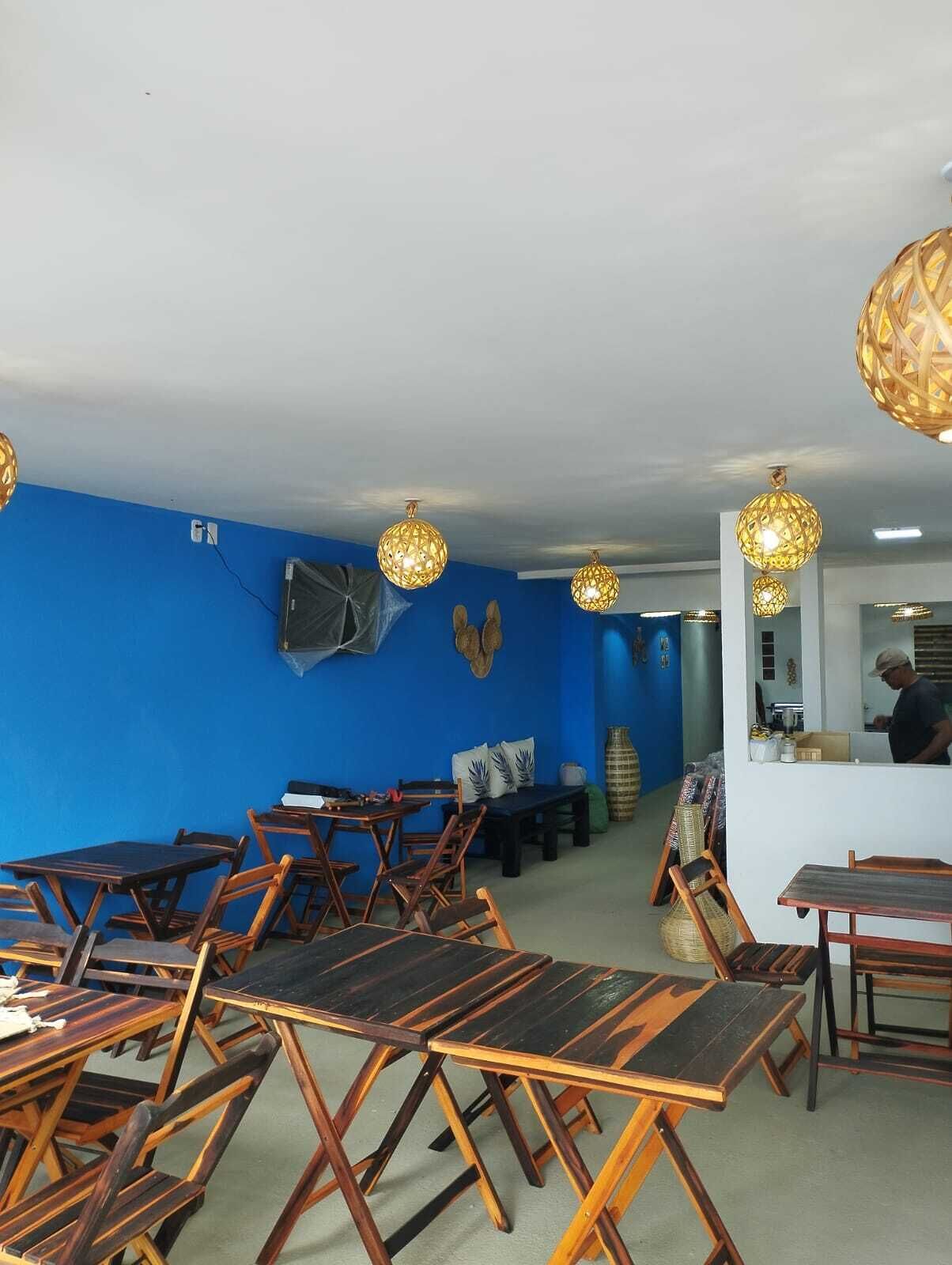 Restaurante com paredes azuis, mesas e cadeiras de madeira e luminárias decorativas penduradas.