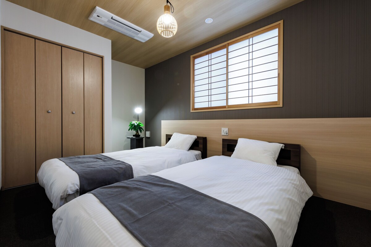 Quarto de hotel com duas camas, um guarda-roupa de madeira, uma janela grande com persianas e um candeeiro de teto.