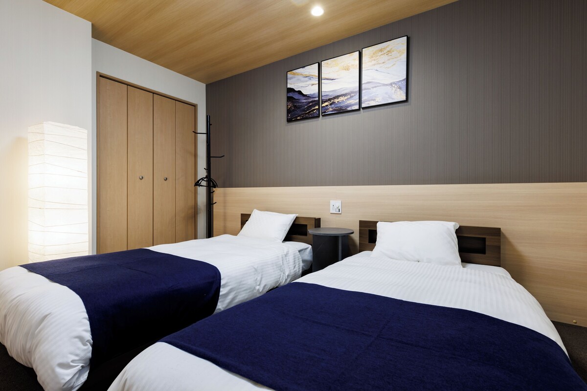 Quarto de hotel com duas camas, um guarda-roupa de madeira, uma mesa redonda pequena e uma obra de arte na parede.