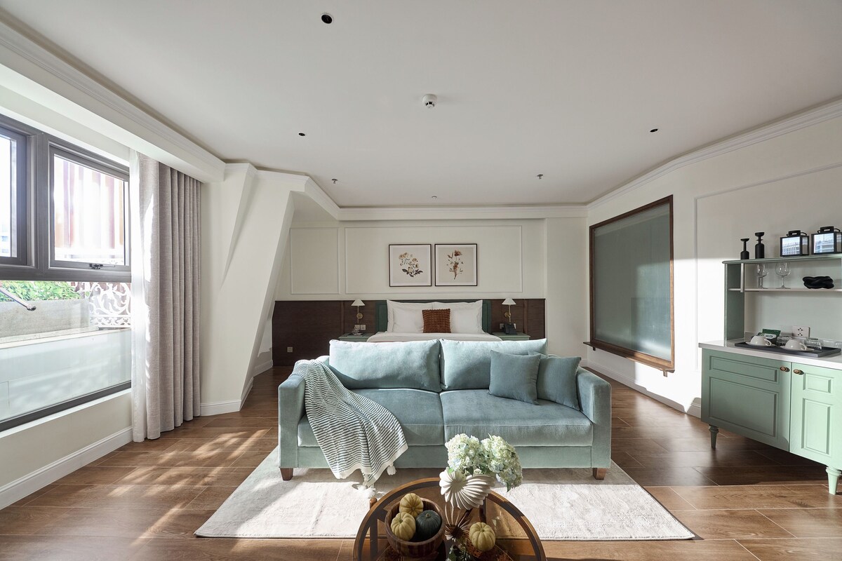 Uma sala de estar moderna com um sofá, uma mesa de centro com objetos decorativos, uma área de cozinha com armários e uma janela grande com cortinas.