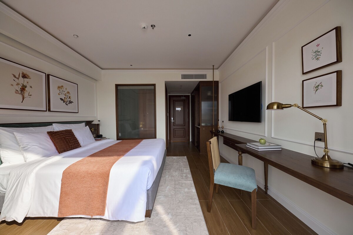 Quarto de hotel com cama, escrivaninha, cadeira, televisão e obras de arte nas paredes.