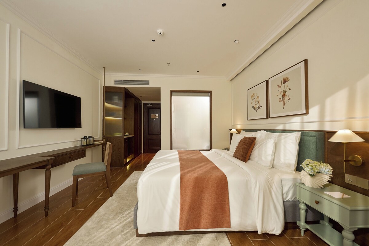 Quarto de hotel com cama, escrivaninha, cadeira, televisão, mesa de cabeceira e obras de arte emolduradas na parede.