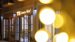 Exterior do TH Luxury Hotel, com placa visível, amplas portas de vidro e um vaso de planta em primeiro plano.