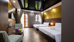 Quarto de hotel com cama, duas mesas de cabeceira, uma cadeira, uma mesinha com objetos e uma janela grande com cortinas.