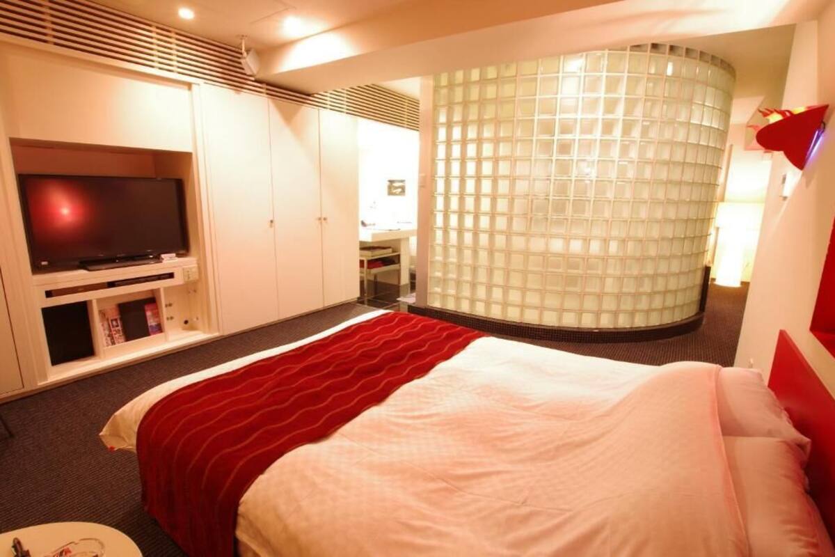 Quarto de hotel com cama, televisão, minibar e banheiro com divisória de vidro.