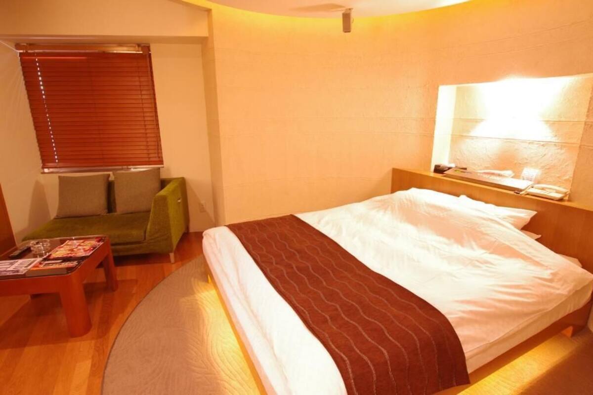 Quarto de hotel com cama, mesa de cabeceira, cadeira, janela com persianas e luminária na parede.