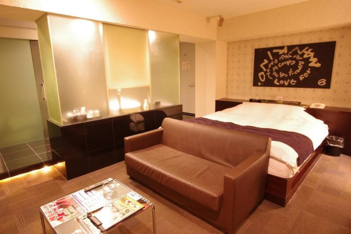 Quarto de hotel com cama, sofá, uma pequena mesa com uma revista, espelho e uma parede com uma frase motivacional.