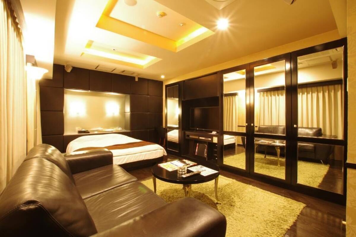 Quarto de hotel moderno com uma cama grande, um sofá, uma mesa de centro e uma porta de vidro deslizante para uma varanda.