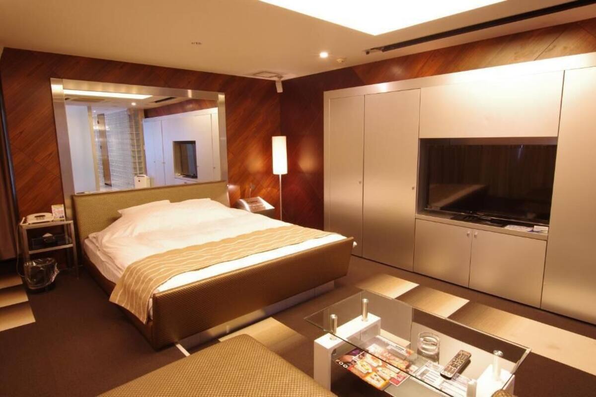 Quarto de hotel com uma cama grande, televisão, espelho e mesa de centro de vidro com controles remotos.