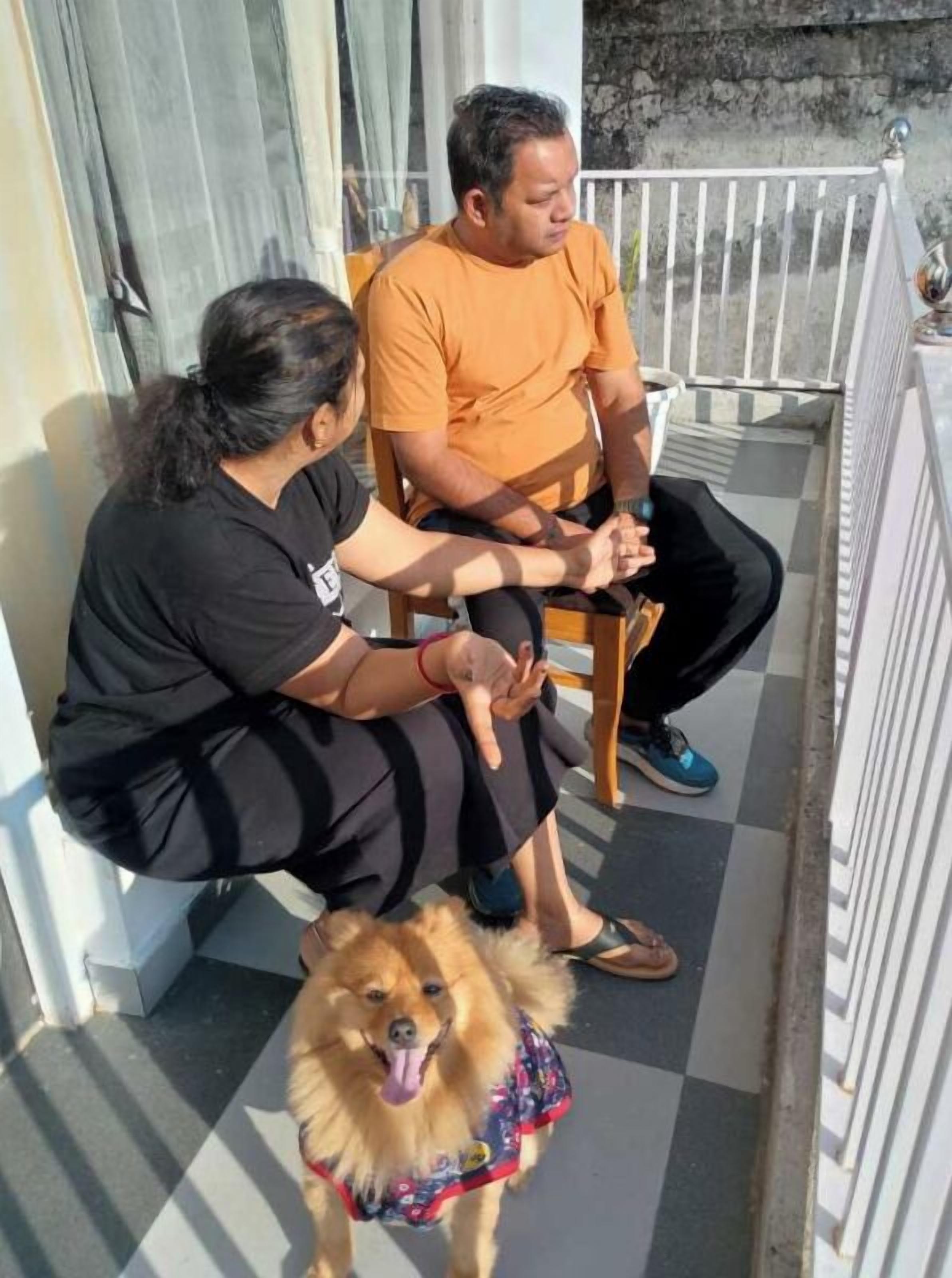 Duas pessoas sentadas num banco numa varanda, com um pequeno cão por perto.