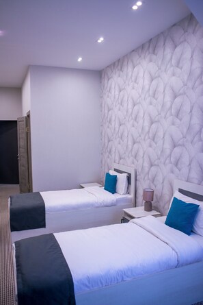 Quarto de hotel com duas camas, parede texturizada, porta e mesa de cabeceira com abajur.