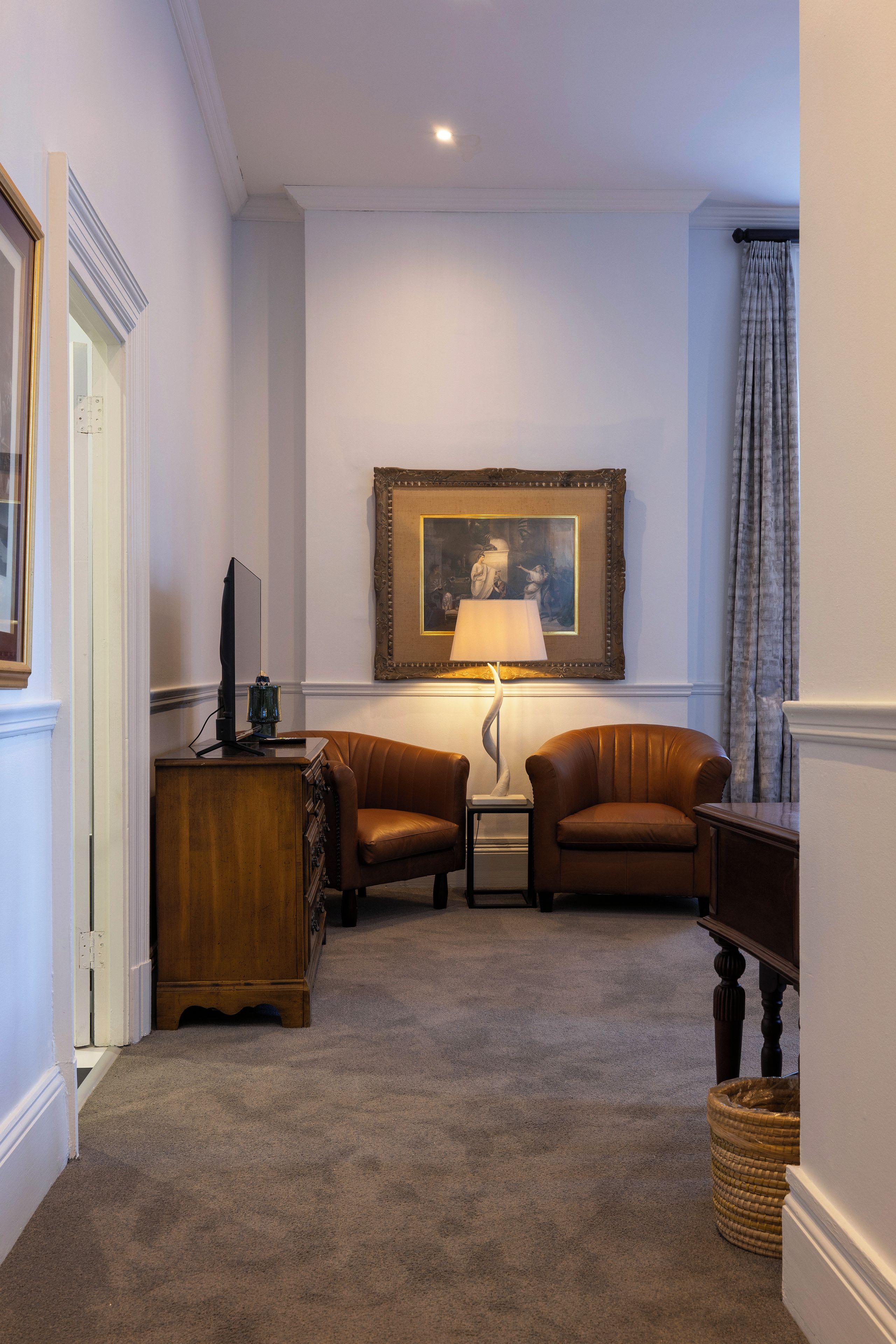 Um quarto com piso carpete, aparador de madeira, duas poltronas de couro, luminária de mesa, obra de arte emoldurada e janela com cortinas.
