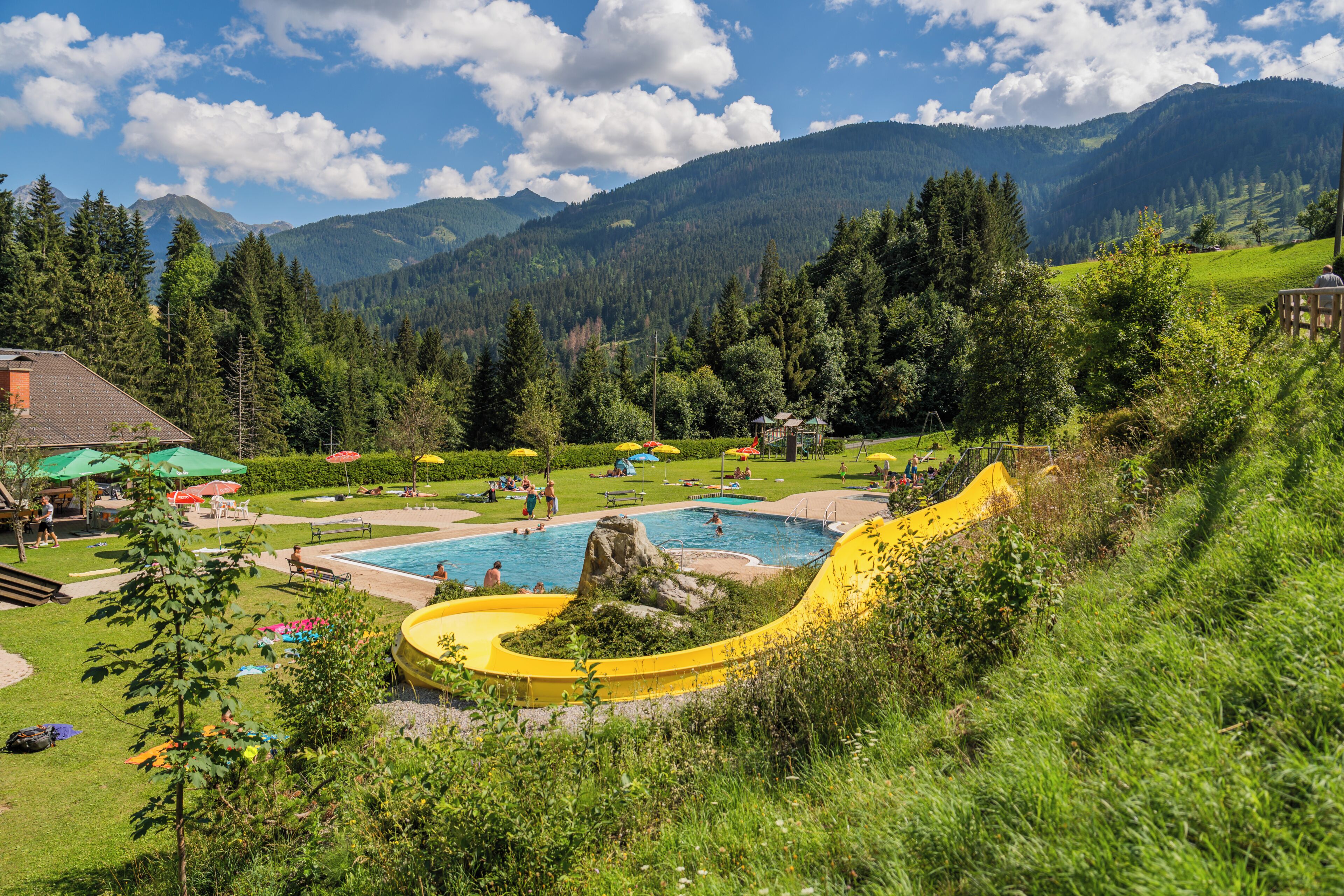Área de piscina com escorrega aquático, cercada por vegetação e montanhas.