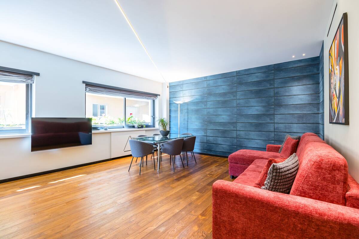 Uma sala de estar moderna com piso de madeira, mesa de jantar com tampo de vidro, sofás vermelhos e uma ampla parede azul decorativa.