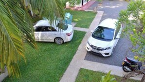 Rua residencial com dois carros estacionados, calçada e uma pequena árvore.