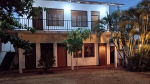Um edifício de dois andares com varanda, escadaria e um pequeno jardim com palmeiras.