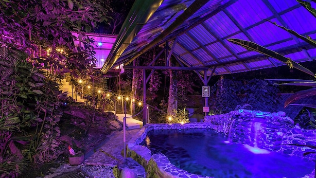 Cena noturna com uma nascente termal, paredes de pedra e uma galeria coberta rodeada por plantas.