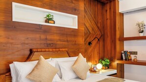 Cama bem arrumada com lençóis brancos e duas almofadas bege, mesa de cabeceira com abajur, uma pequena planta e prateleira com livros e um relógio.