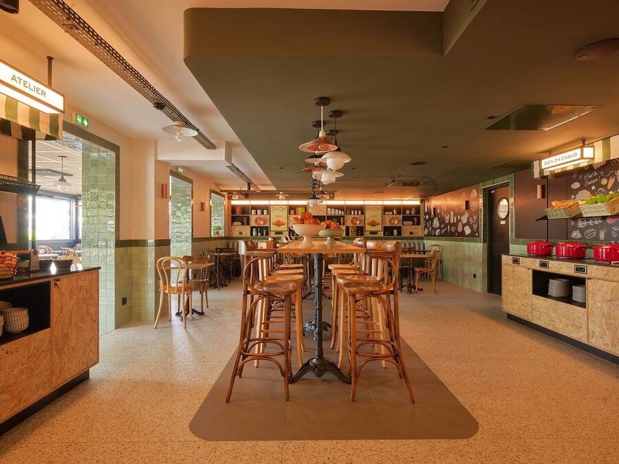Interior de restaurante com uma mesa central rodeada por cadeiras, área de cozinha com panelas vermelhas e um balcão exibindo diversos alimentos.