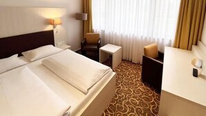 Quarto de hotel com cama, escrivaninha, cadeira, luminária e janela com cortinas.