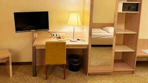 Quarto de hotel compacto com escrivaninha, cadeira, televisão, abajur e cama com guarda-roupa.