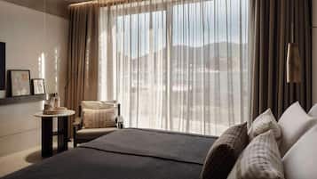 Quarto de hotel moderno com cama grande, mesinha de cabeceira, uma cadeira e uma janela ampla com cortinas.