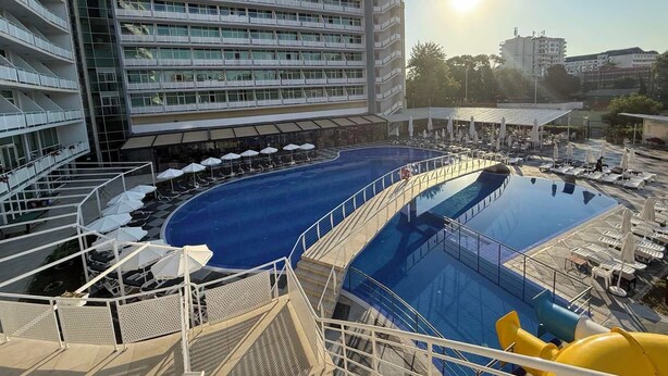 Piscina com áreas para banho de sol, um edifício com vários andares e céu claro.