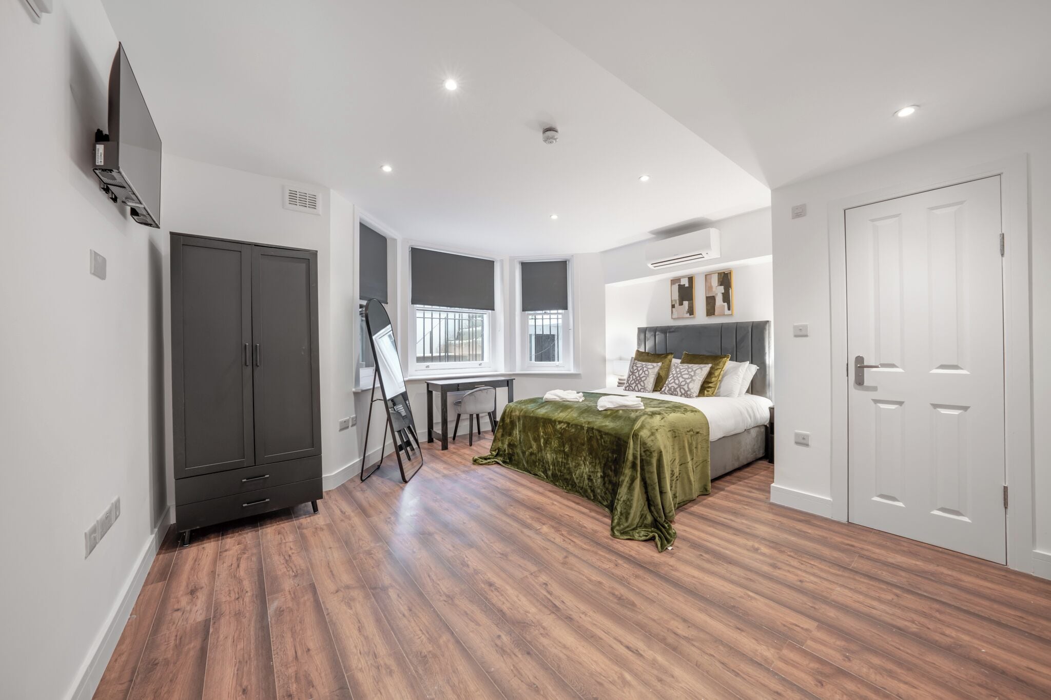 Quarto com cama, escrivaninha, guarda-roupa e TV fixada na parede.
