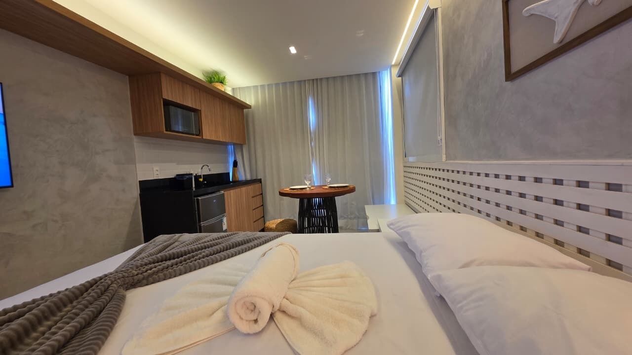 Quarto de hotel com cama, uma pequena mesa com uma toalha, cozinha compacta e janela com cortinas.