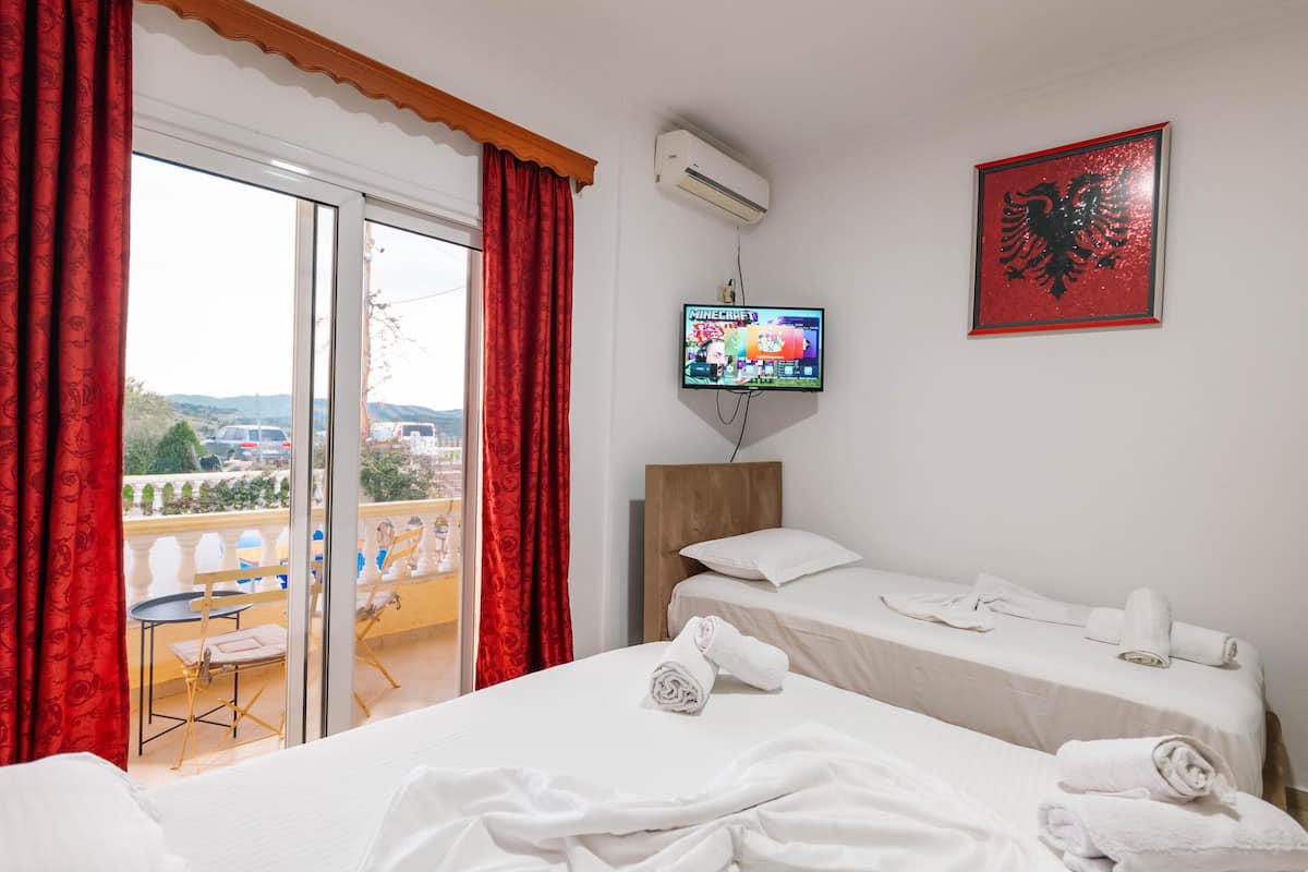 Quarto de hotel com duas camas, televisão, varanda com cortinas vermelhas e ar condicionado fixado na parede.