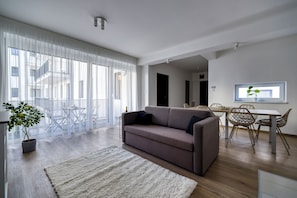 Uma sala de estar moderna com sofá, mesa de jantar com cadeiras, planta em vaso e grandes janelas.