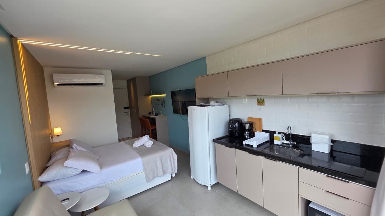Quarto de hotel compacto com uma cama, uma pequena mesa de escritório, uma cozinha compacta com geladeira, cafeteira e pia.
