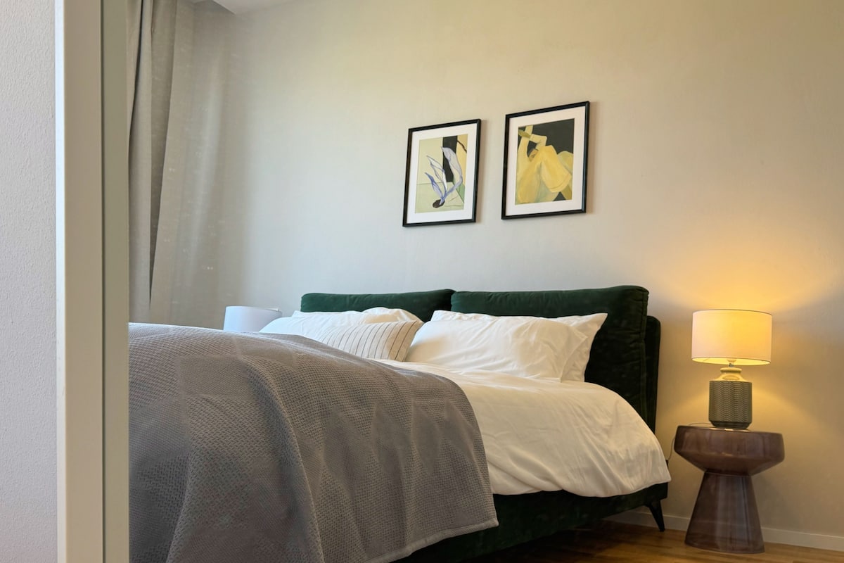 Uma cama bem arrumada com cabeceira verde, uma luminária de cabeceira e duas obras de arte emolduradas na parede.