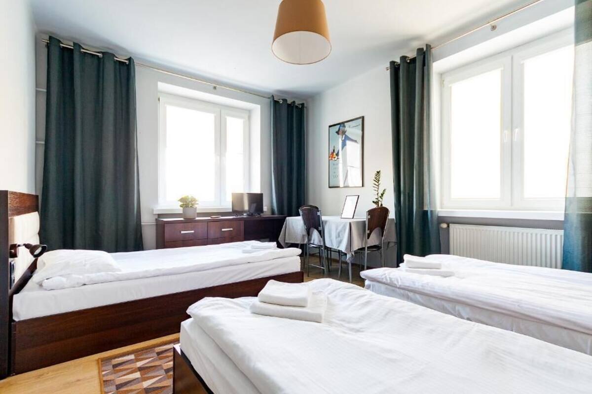 Quarto de hotel com duas camas de solteiro, uma escrivaninha, uma cadeira, uma mesinha, uma janela com cortinas e um radiador.