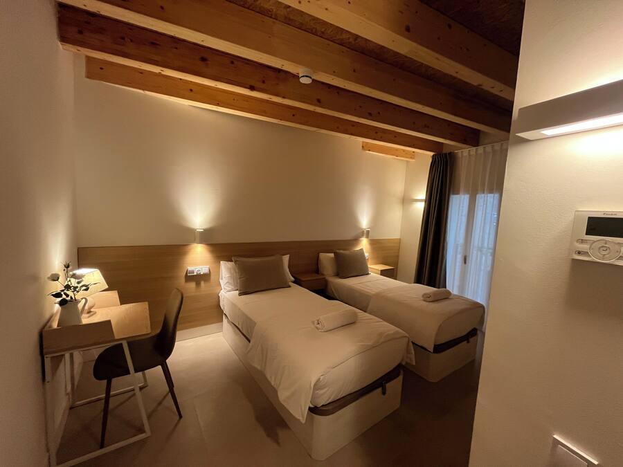 Quarto de hotel com duas camas, uma escrivaninha, uma cadeira e um termostato na parede.