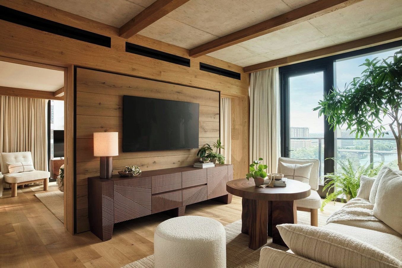 Uma sala de estar moderna com uma grande TV de tela plana, painéis de madeira, uma mesa de centro redonda e vista para um horizonte urbano através de amplas janelas.