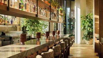 Uma área de bar com prateleiras de garrafas, banquetas e plantas em vasos.