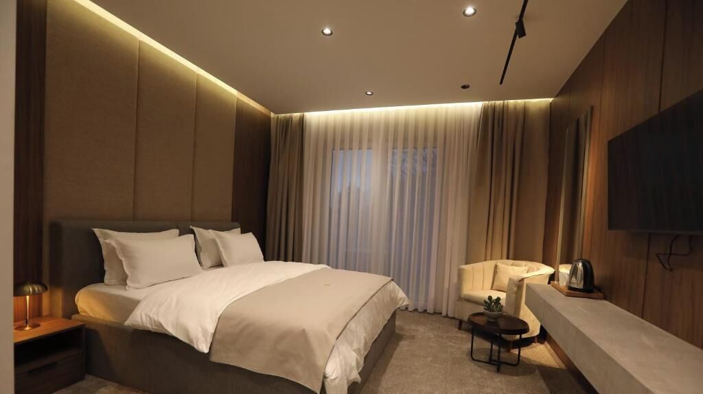 Quarto de hotel moderno com uma cama grande, área de estar com uma cadeira e mesa, televisão de tela plana e uma janela ampla com cortinas.