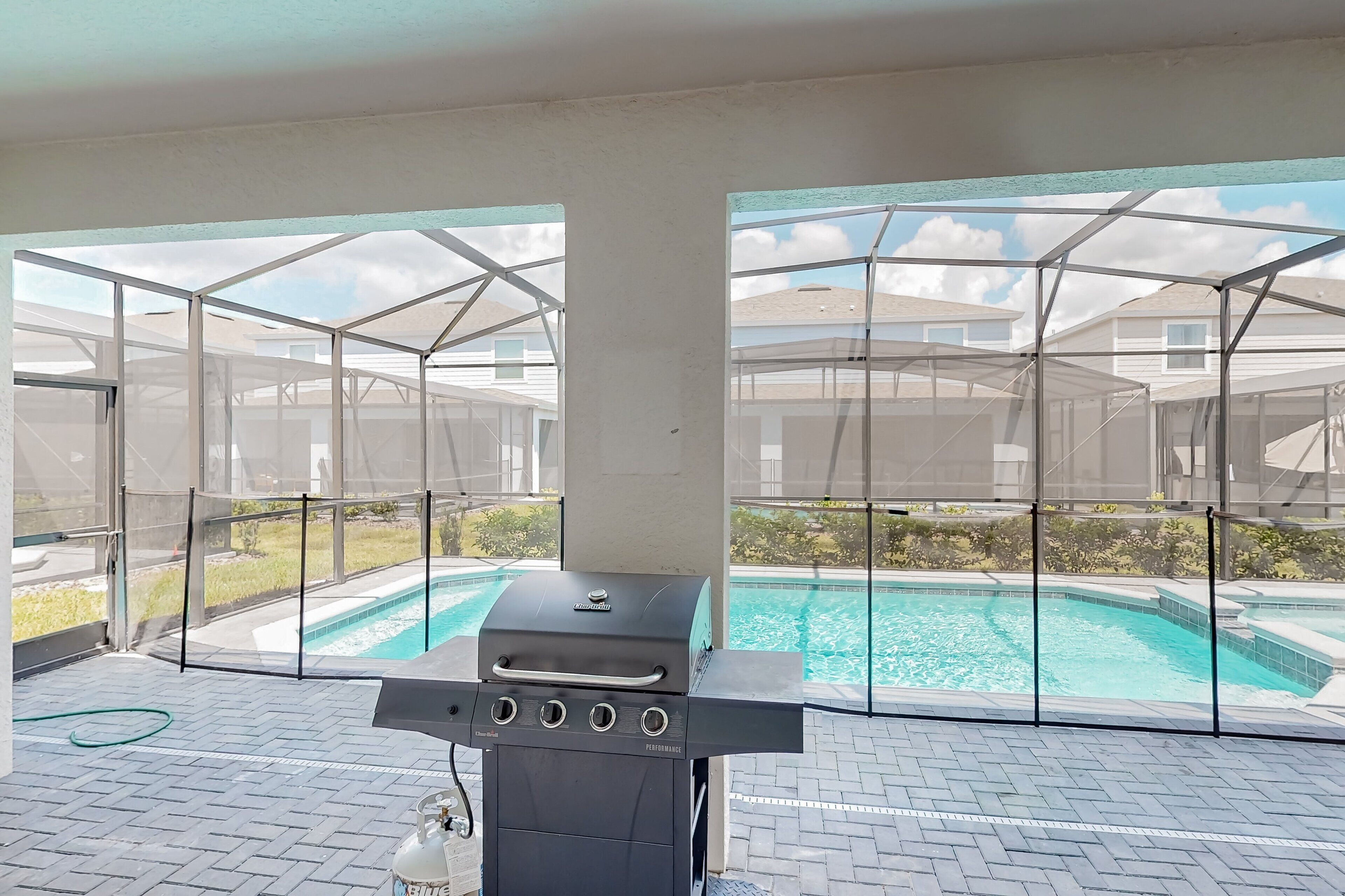 Área externa coberta com churrasqueira, piscina e vista para casas através de uma proteção.